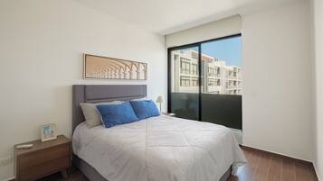 Quarto moderno com uma cama grande, uma mesinha de cabeceira, um quadro emoldurado e vista para edifícios através de uma janela ampla.