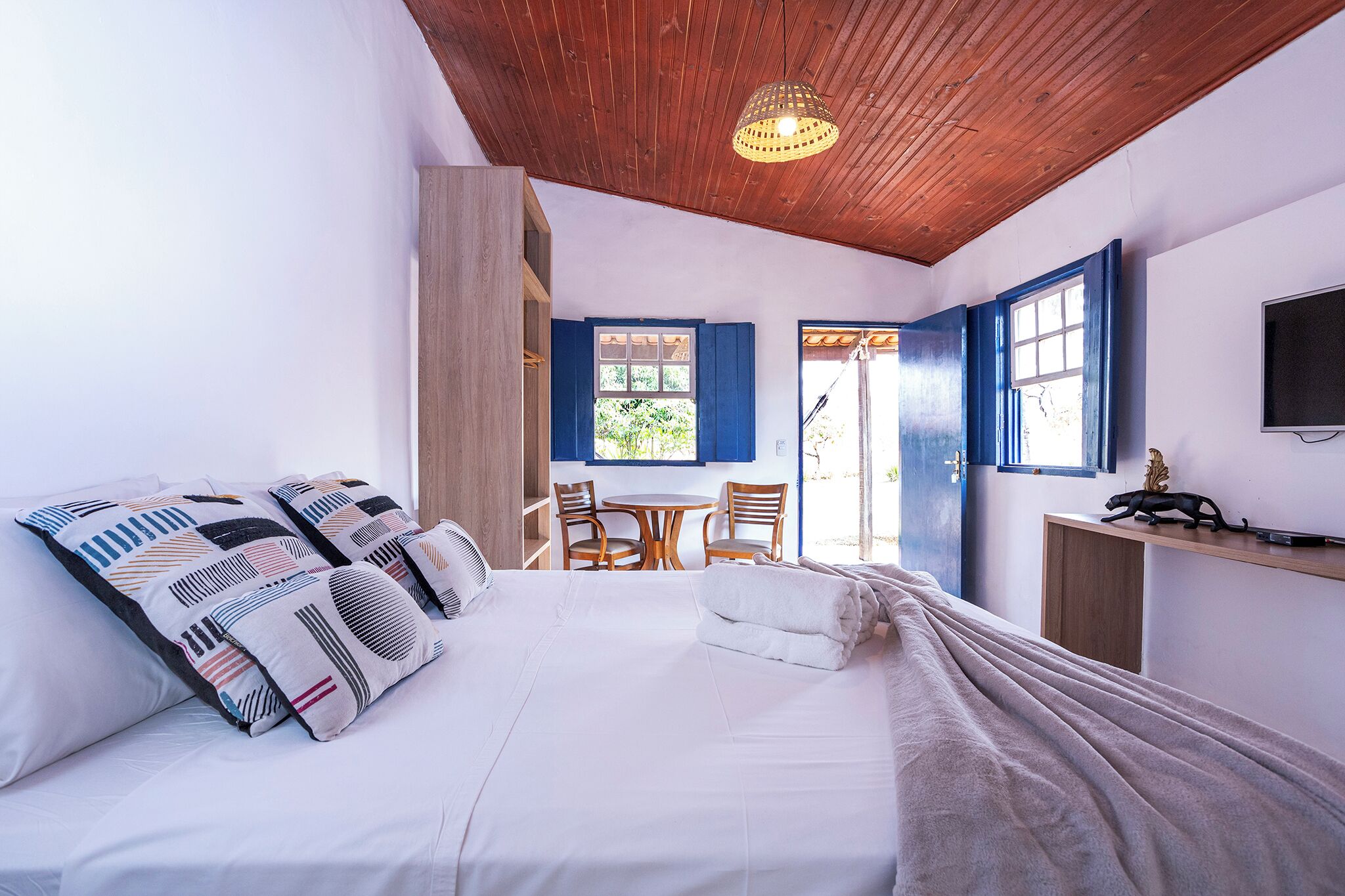 Quarto com cama, escrivaninha, cadeiras, televisão e janela.
