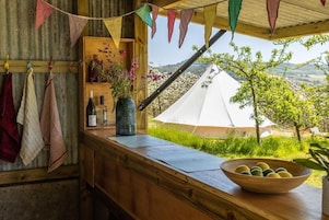 Uma cabana rústica de madeira com um banco de madeira, uma prateleira de madeira com garrafas e um vaso de flores, e vista para uma tenda branca e vegetação.