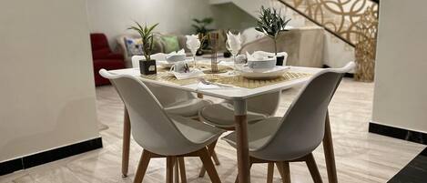 Uma sala de jantar com uma mesa posta para dois, cadeiras brancas e uma escada ao fundo.