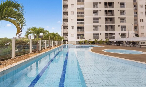 Piscina com deck ladrilhado, cercada por uma grade e palmeiras.