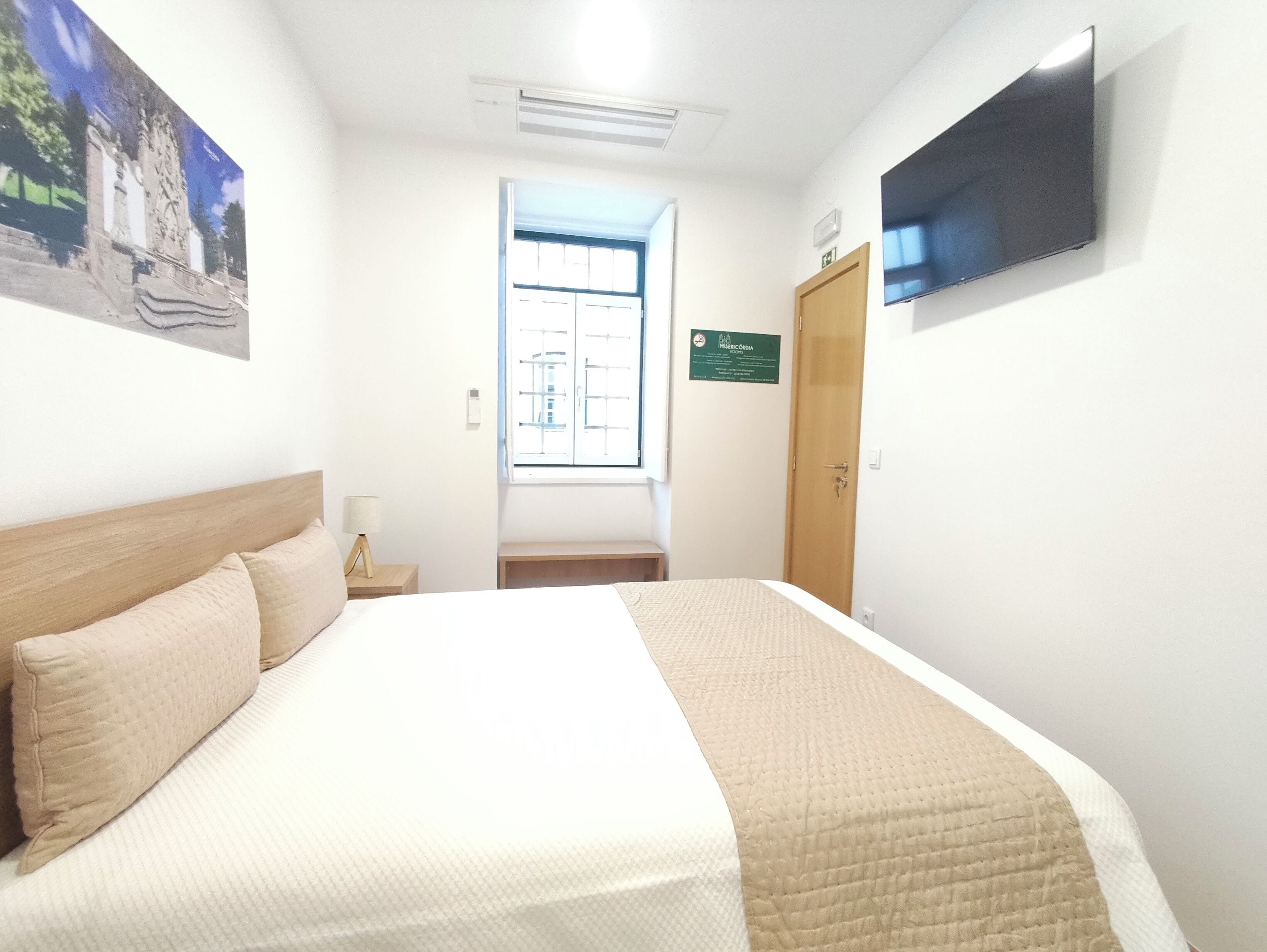 Quarto de hotel com cama, televisão fixada na parede, janela e porta.