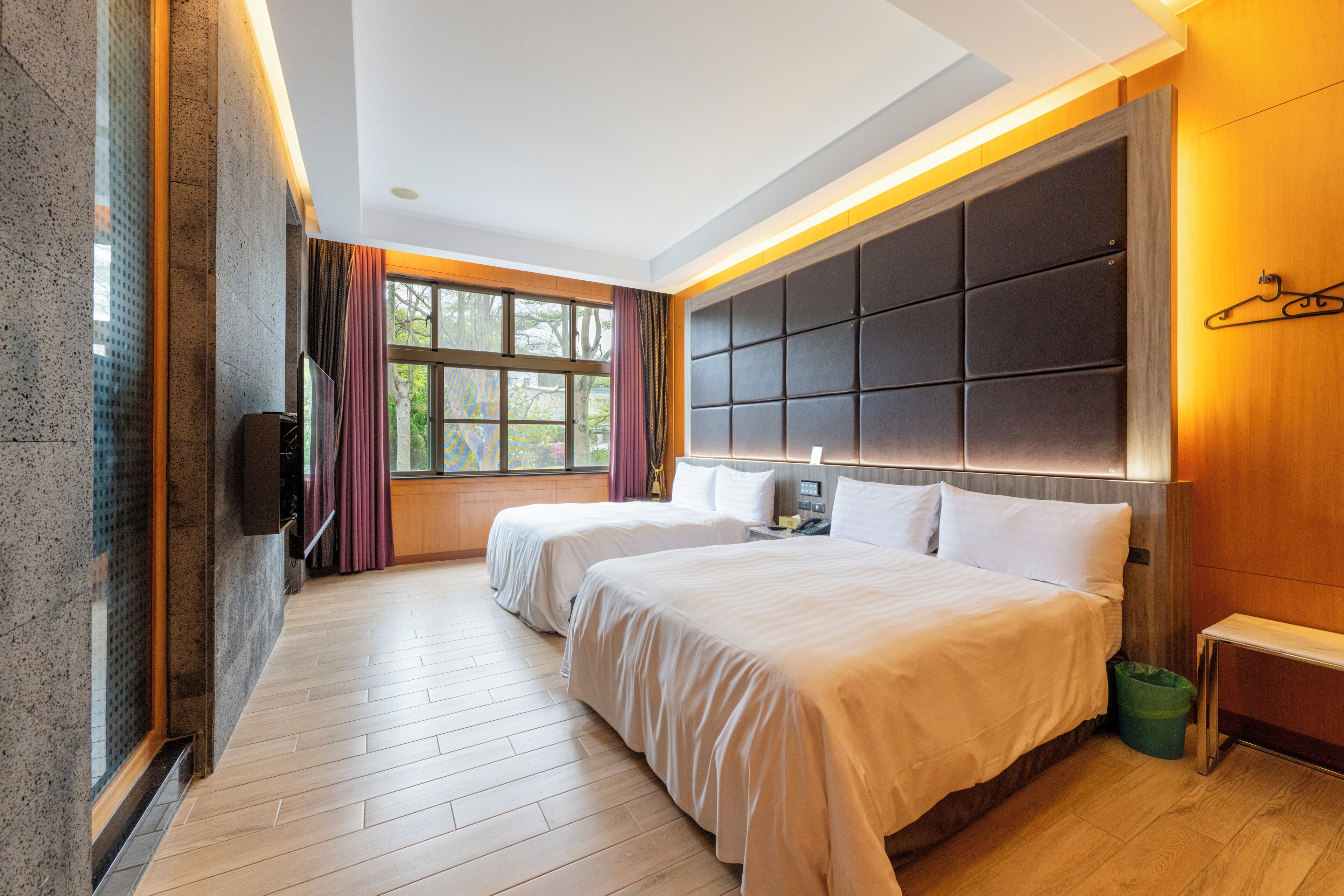 Quarto de hotel com duas camas, uma janela grande com cortinas, televisão na parede e piso de madeira.