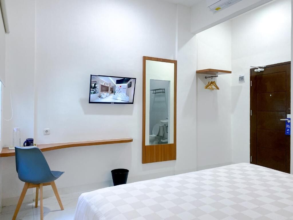 Quarto de hotel com uma cama, uma escrivaninha com cadeira, televisão, espelho, prateleira de madeira e uma porta.