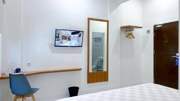 Quarto de hotel com uma cama, uma escrivaninha com cadeira, televisão, espelho, prateleira de madeira e uma porta.
