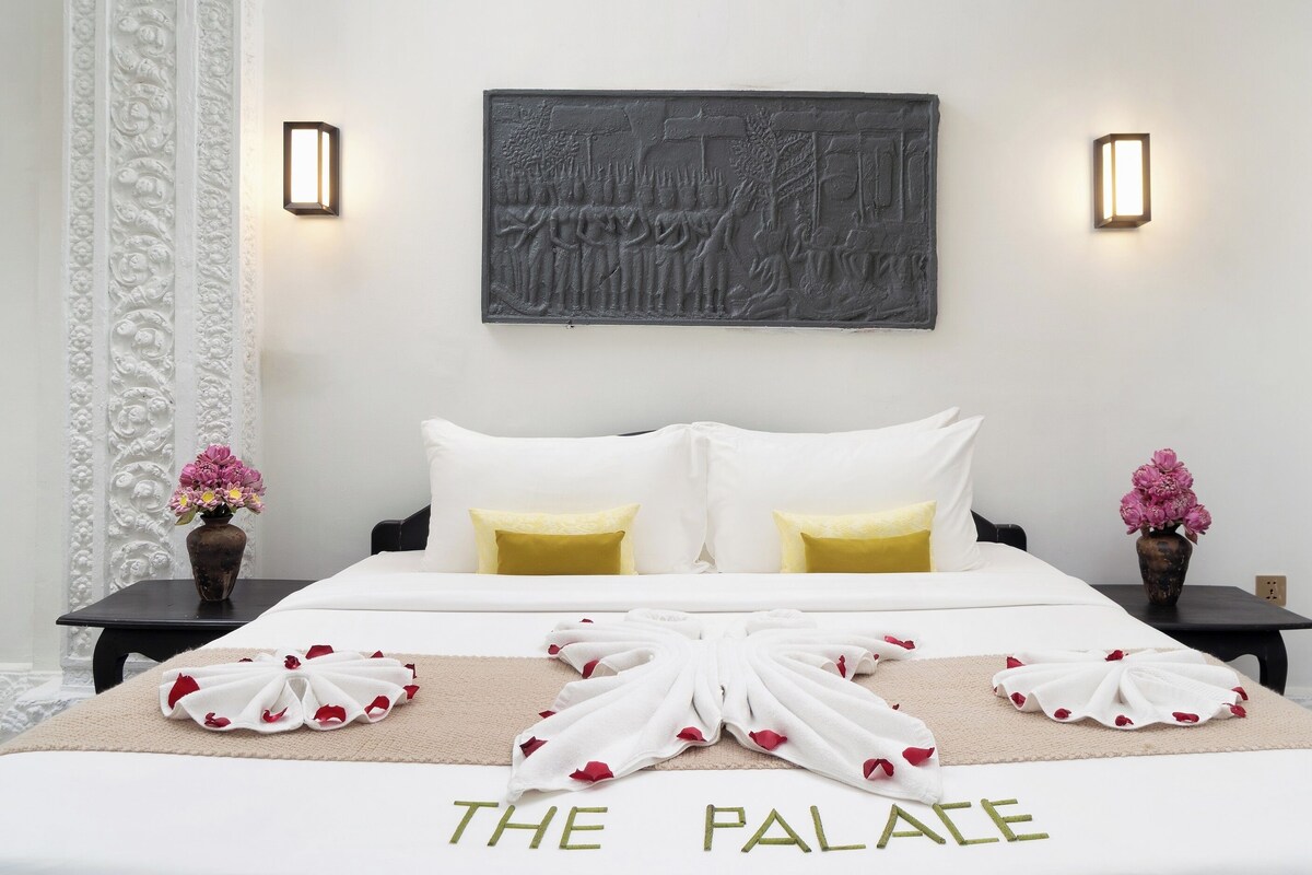 Quarto de hotel com cama, duas mesas de cabeceira, um objeto decorativo na parede e uma placa na cama com a inscrição 'THE PALACE'.