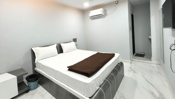Quarto de hotel moderno com cama, ventilador de teto, iluminação embutida, ar condicionado, uma pequena lixeira e banheiro com vaso sanitário e pia.