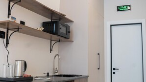 Cozinha moderna com micro-ondas, máquina de café e pia. Há uma prateleira de madeira acima do balcão e uma porta com uma placa indicando um número de quarto.