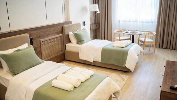 Quarto de hotel com duas camas, uma mesinha com cadeiras, uma luminária e vista para o exterior.