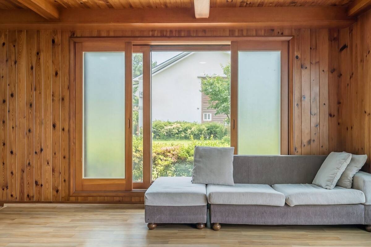 Quarto com paredes de madeira, sofá cinza e porta de vidro deslizante para um jardim.