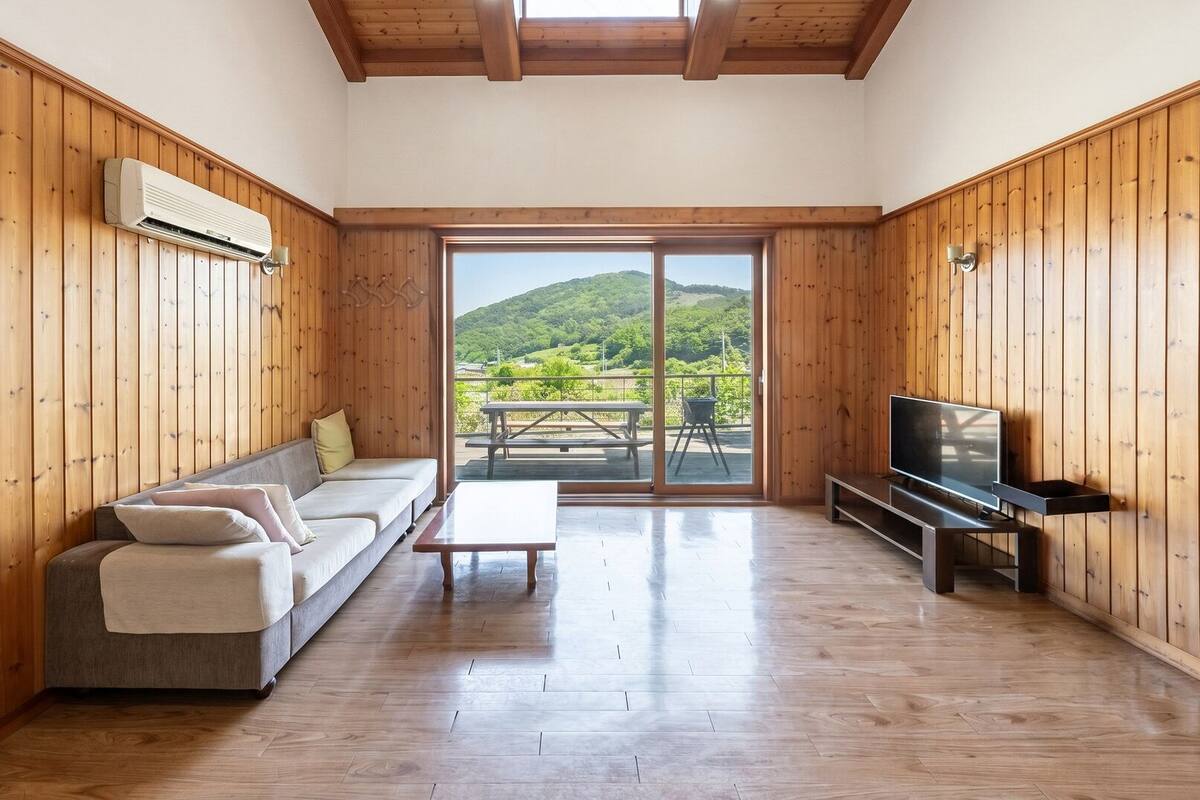 Quarto com paredes de madeira, um sofá, uma televisão e vista para uma paisagem verde através de uma porta deslizante.