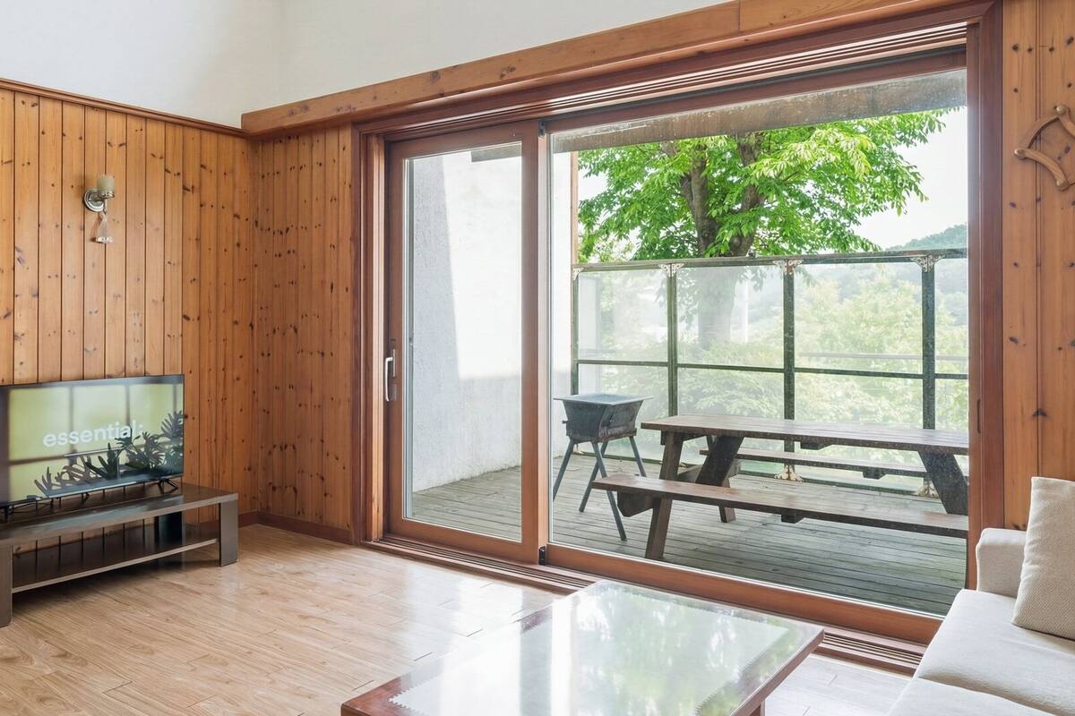 Quarto com paredes de madeira, uma porta de vidro deslizante para um deck ao ar livre, uma televisão em um suporte e uma mesa de centro de vidro.