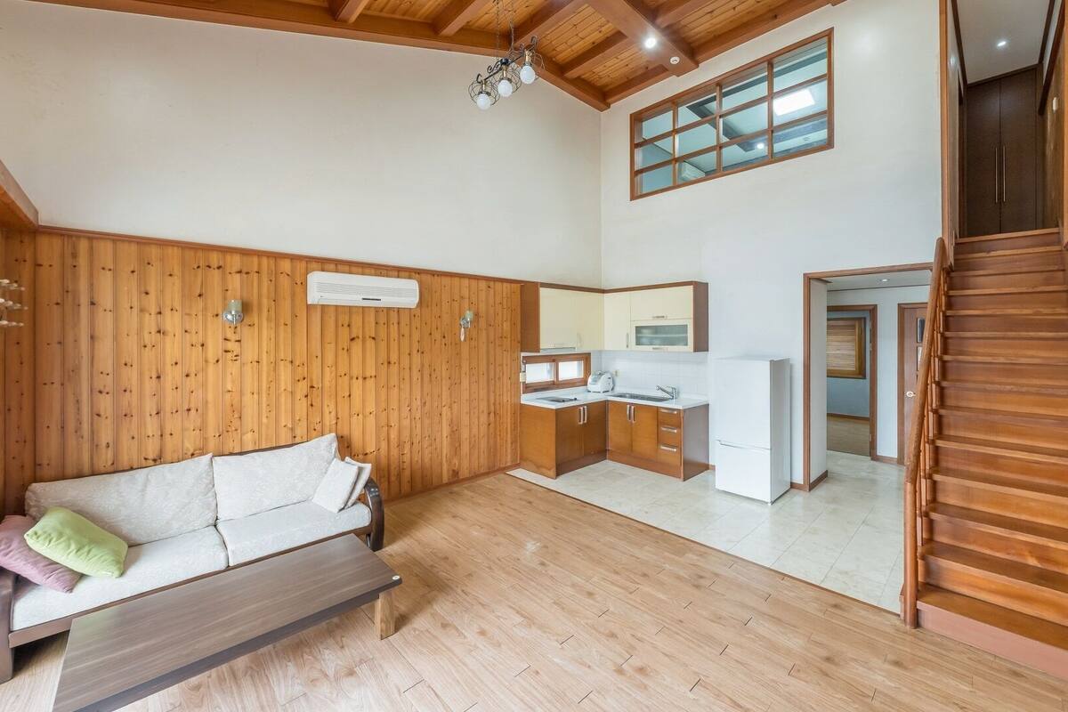 Um quarto amplo com piso de madeira, um sofá, área de cozinha com geladeira e uma escada.