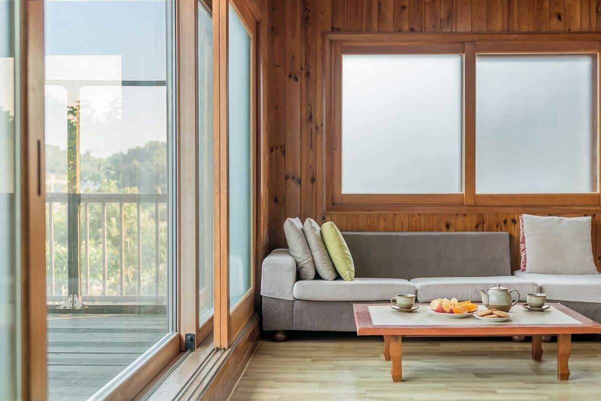 Um quarto com parede revestida em madeira, um sofá cinza com almofadas, uma mesa de centro de madeira com bule e xícaras, e vista através de grandes janelas.