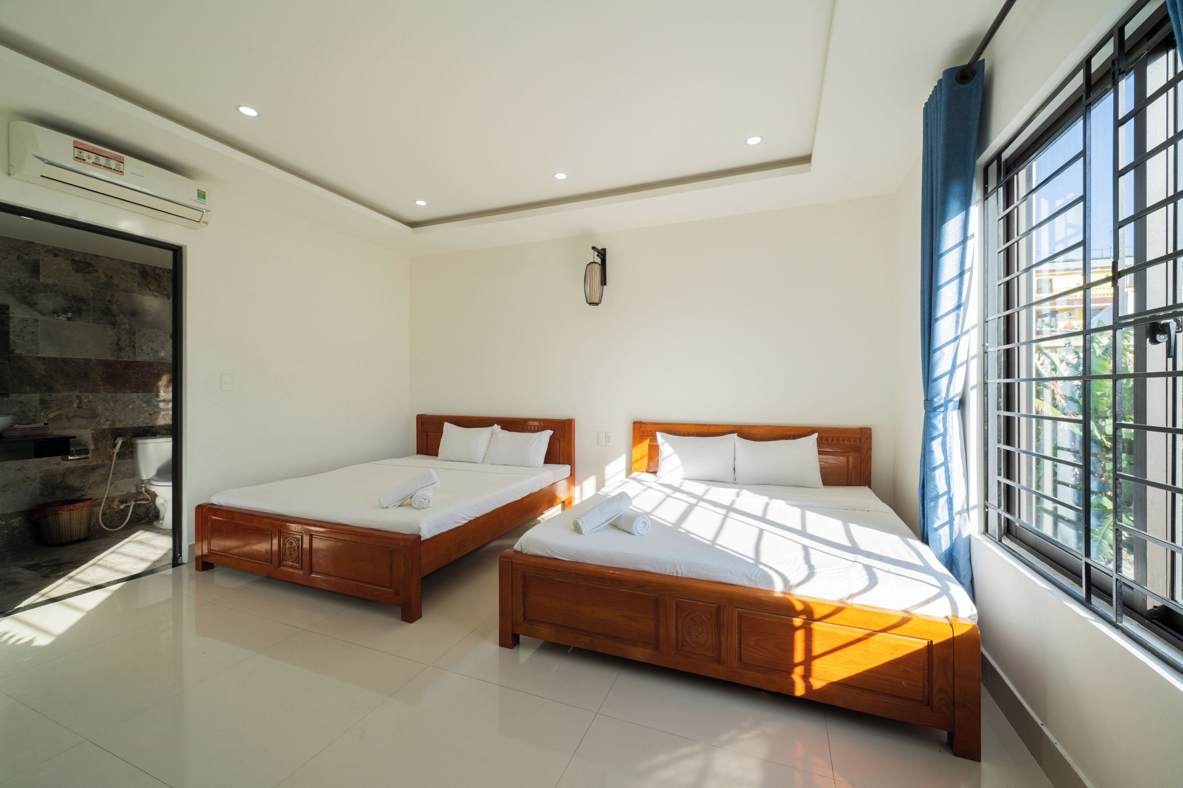 Quarto de hotel com duas camas de madeira, parede de pedra, janela com grades de metal e ar condicionado instalado no teto.