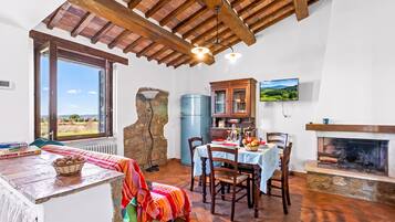 Cozinha rústica com parede de pedra, vigas de madeira no teto, mesa de jantar posta para quatro pessoas e lareira.