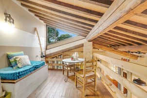 Quarto em estilo loft com teto de madeira, cama com roupa de cama azul, uma mesinha com cadeiras e janela com vista para árvores.