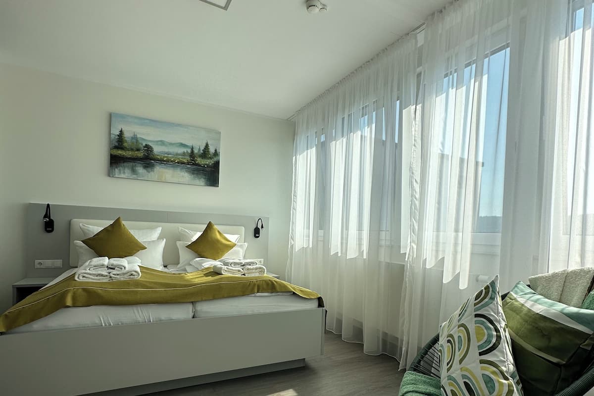 Quarto moderno com cama, uma cadeira, um quadro e cortinas leves.