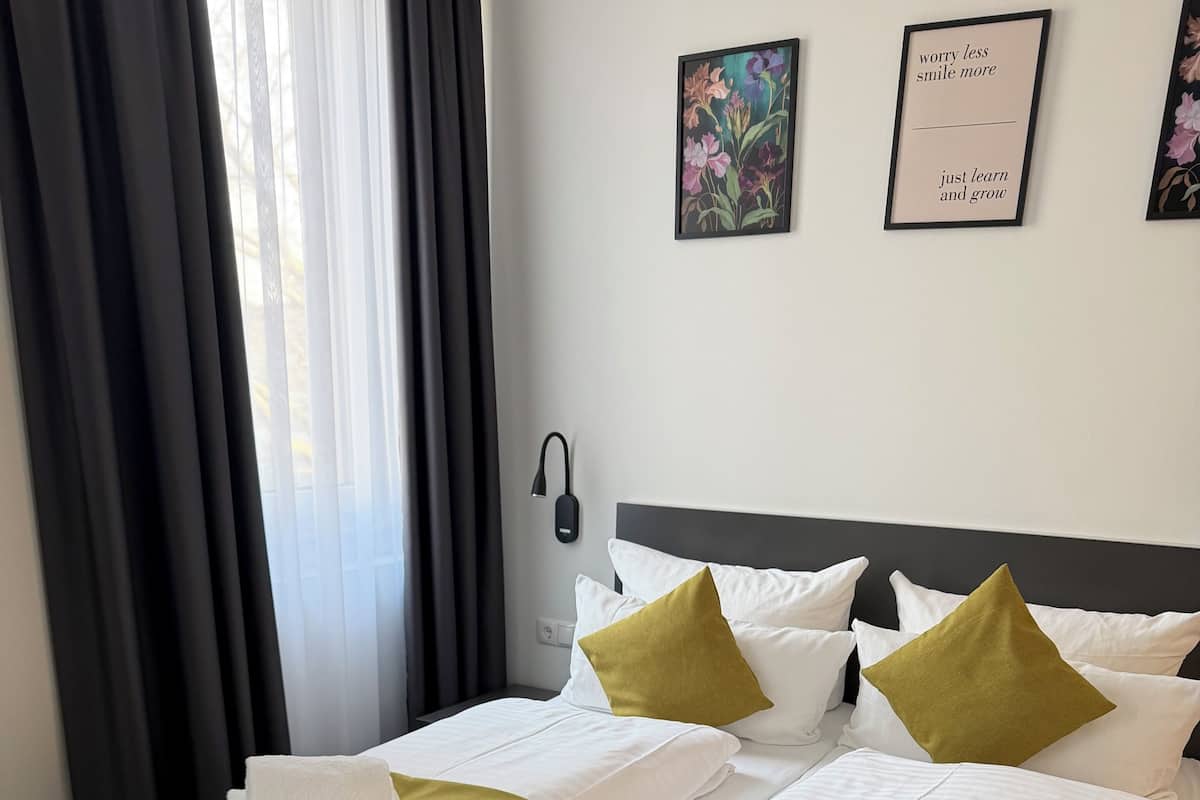 Cama bem arrumada com roupa de cama branca e verde oliva, um pequeno objeto decorativo e uma citação inspiradora emoldurada na parede.