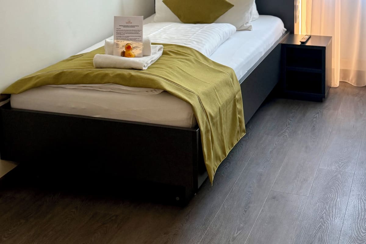 Uma cama bem arrumada com cabeceira escura, mesa de cabeceira com abajur e um quadro na parede.