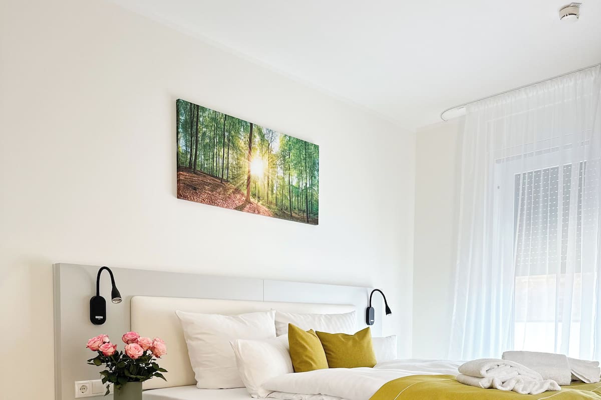 Quarto moderno com cama, mesas de cabeceira, luminária de parede, um quadro e janela com cortinas translúcidas.