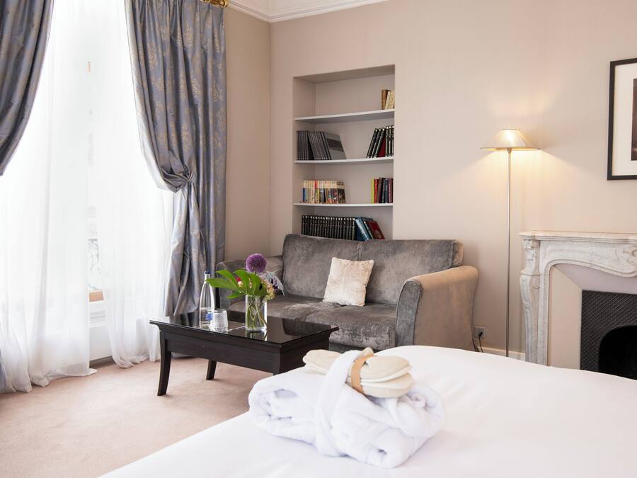 Quarto de hotel com cama, sofá cinzento, estante de livros, lareira e mesa de centro com um vaso e flores.