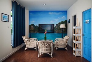 Quarto com vista para o mar azul e falésias, uma mesa redonda com cadeiras, uma televisão e uma porta azul.