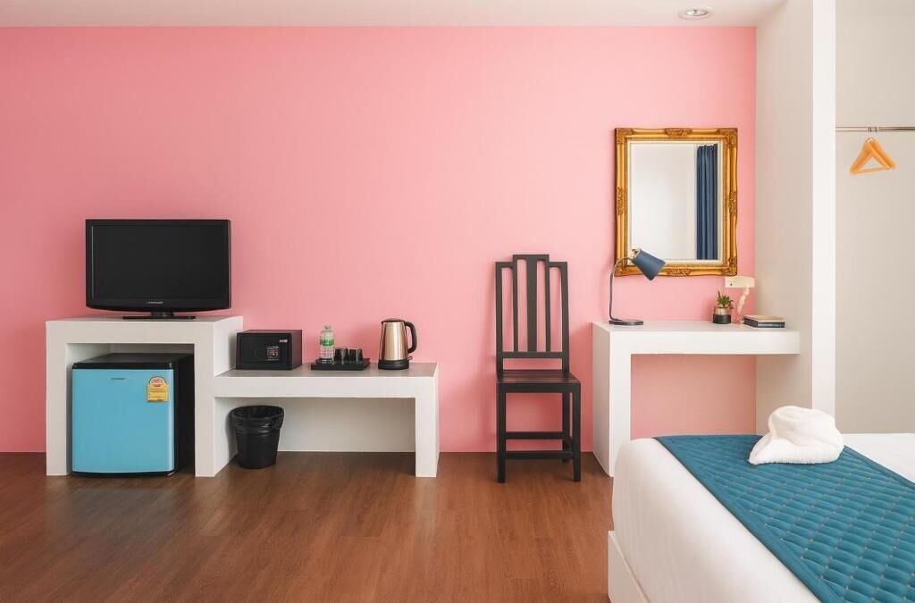 Quarto de hotel com parede rosa, cama com colcha azul, escrivaninha com computador, televisão, pequeno frigorífico azul e uma cadeira.