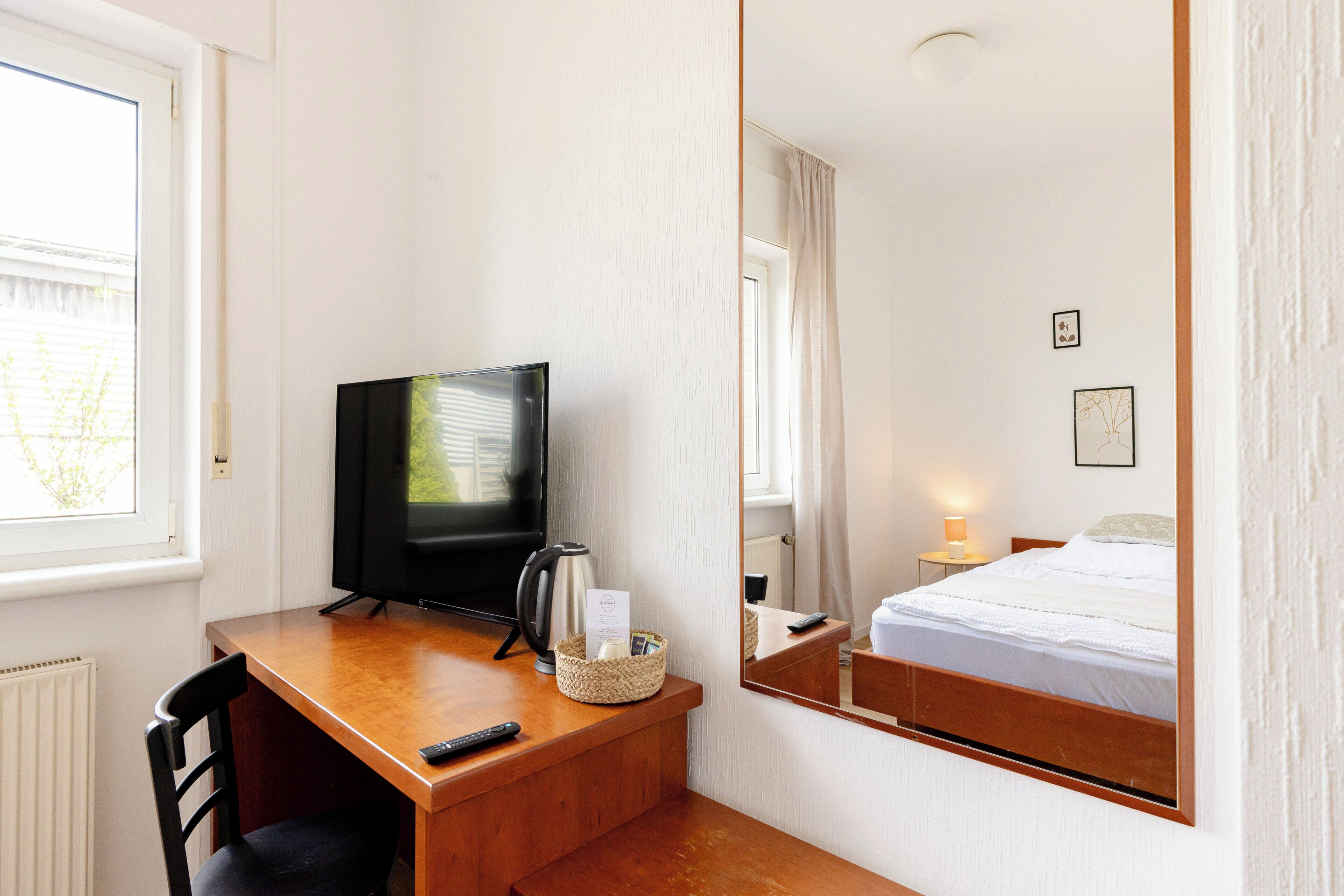 Quarto de hotel com escrivaninha de madeira, televisão, mesa de cabeceira com abajur, cama e um espelho grande.