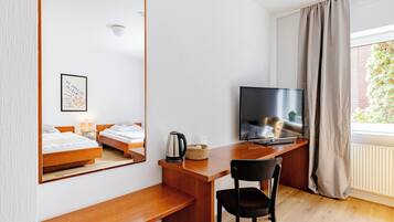 Quarto de hotel com cama, secretária com televisão, cadeira, janela com cortinas e radiador.