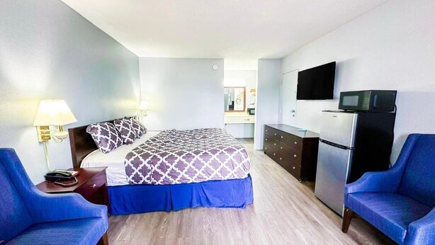 Quarto de hotel com cama, duas cadeiras azuis, uma escrivaninha com abajur, televisão e kitchenette.