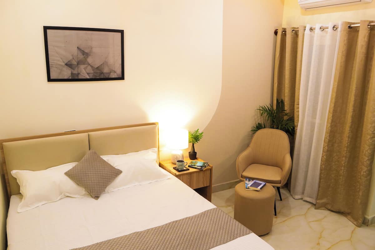 Quarto de hotel com cama, uma cadeira, mesa de cabeceira, abajur, quadro pendurado na parede e cortina.