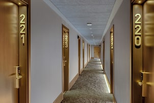 Corredor de hotel com portas numeradas de 221 a 225, piso coberto de carpete e teto com iluminação embutida.