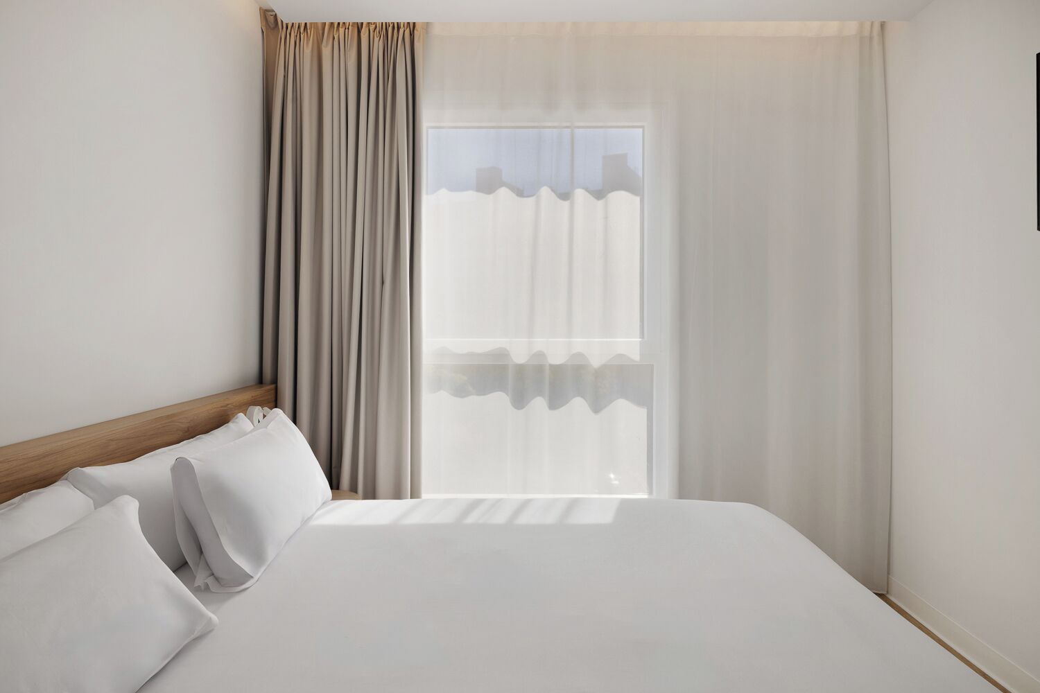 Quarto de hotel com uma cama grande, cabeceira de madeira e cortinas finas.