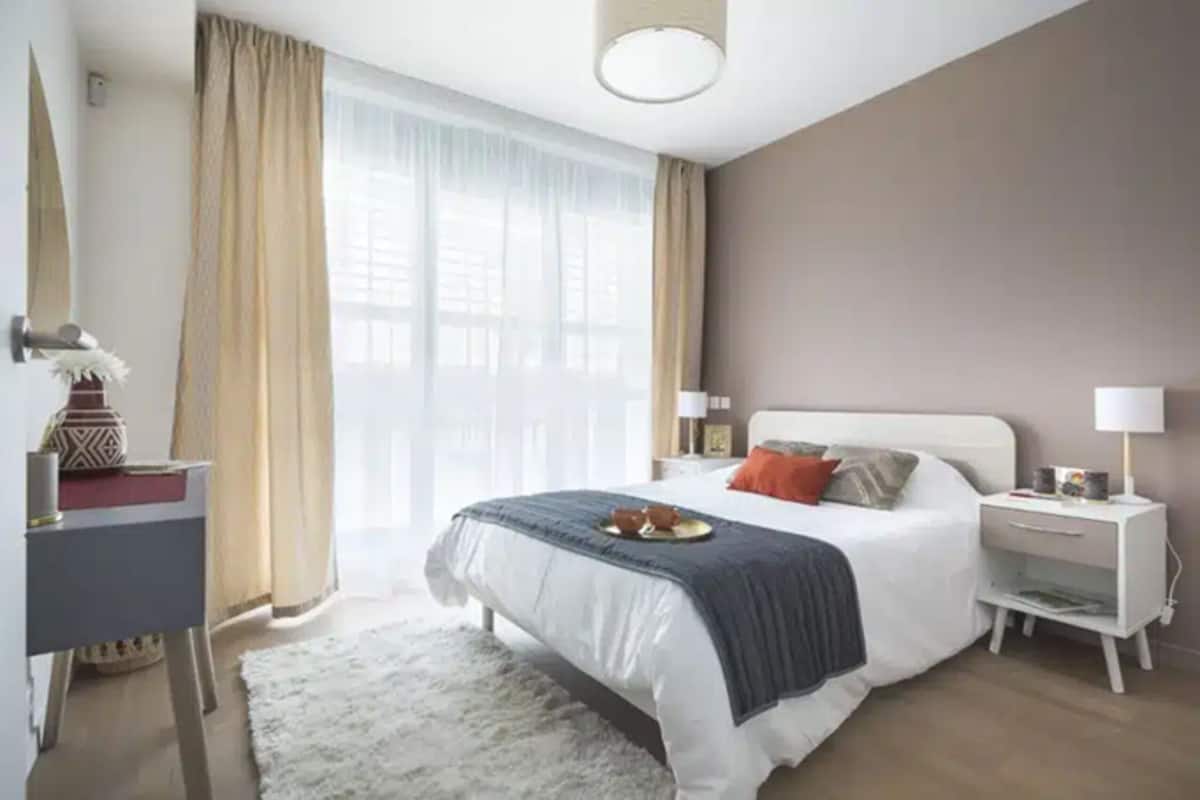 Quarto com cama, mesas de cabeceira, escrivaninha, luminária e janela com cortinas.