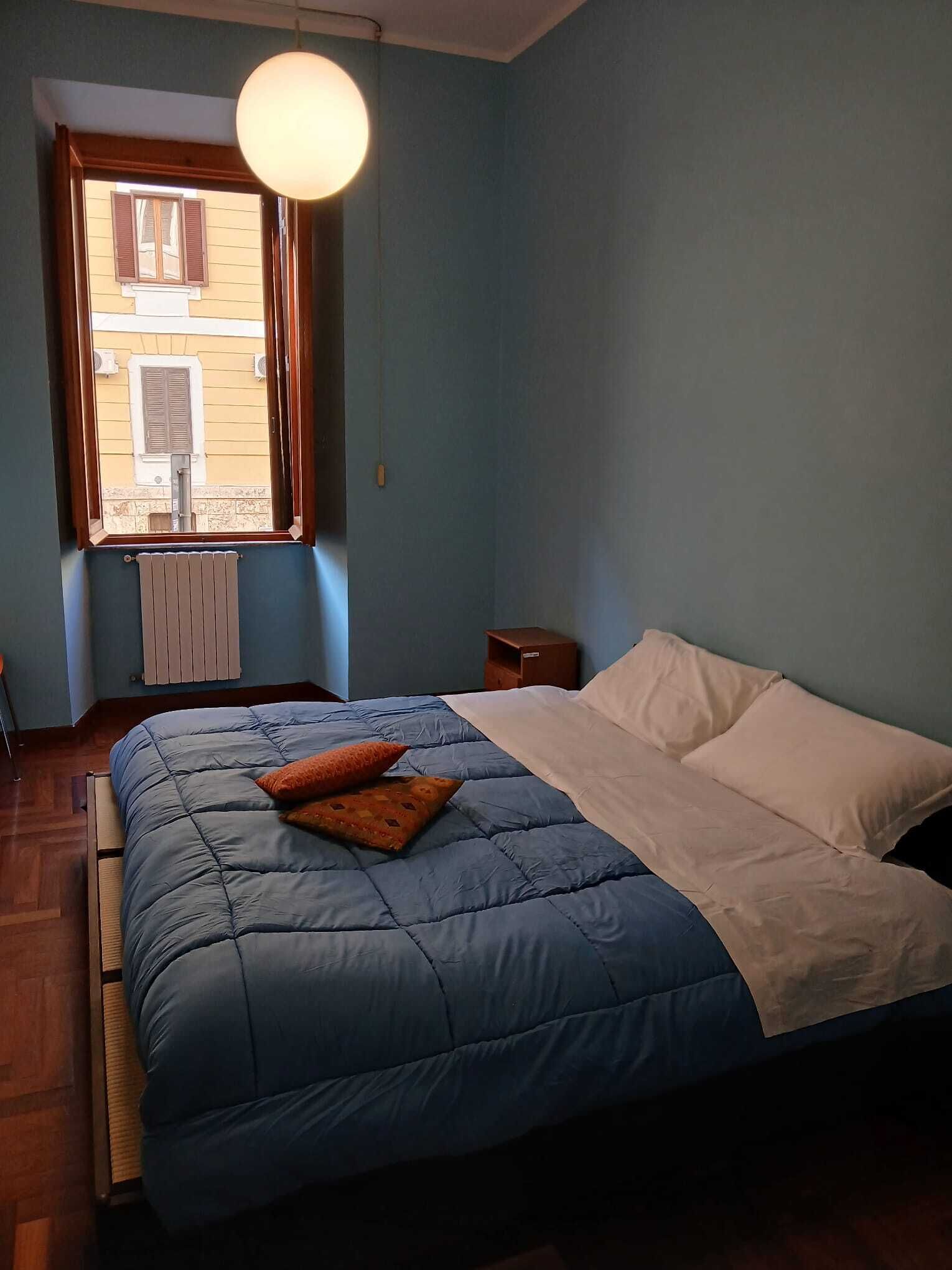 Quarto com cama, janela com persianas, radiador e luminária pendente.