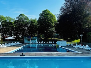 Piscina com espreguiçadeiras, um pequeno edifício e árvores ao fundo.