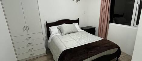 Um quarto com cama, guarda-roupa, mesa de cabeceira, janela com cortinas e luz no teto.