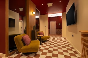 Um hall de hotel com piso xadrez, dois poltronas com almofadas, uma TV de tela plana na parede e uma porta com a inscrição 'ROOM 09-13'.