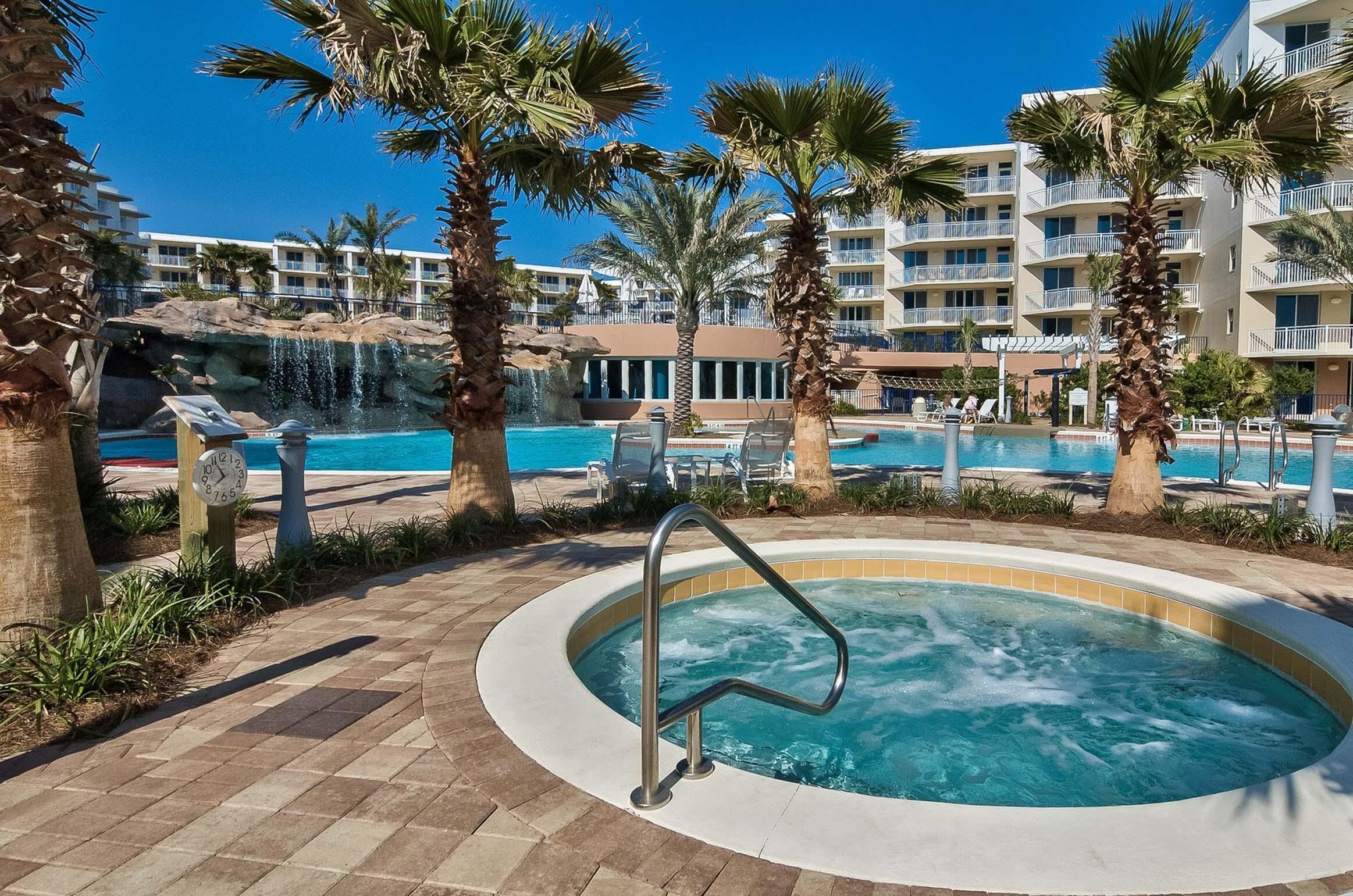 Área de piscina de hotel com hidromassagem, palmeiras e uma cascata.
