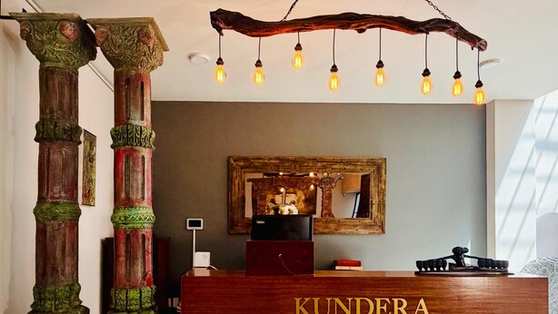 Bancada de recepção com a sinalização 'KUNDERA', um espelho e teto de madeira rústica com luminárias pendentes.