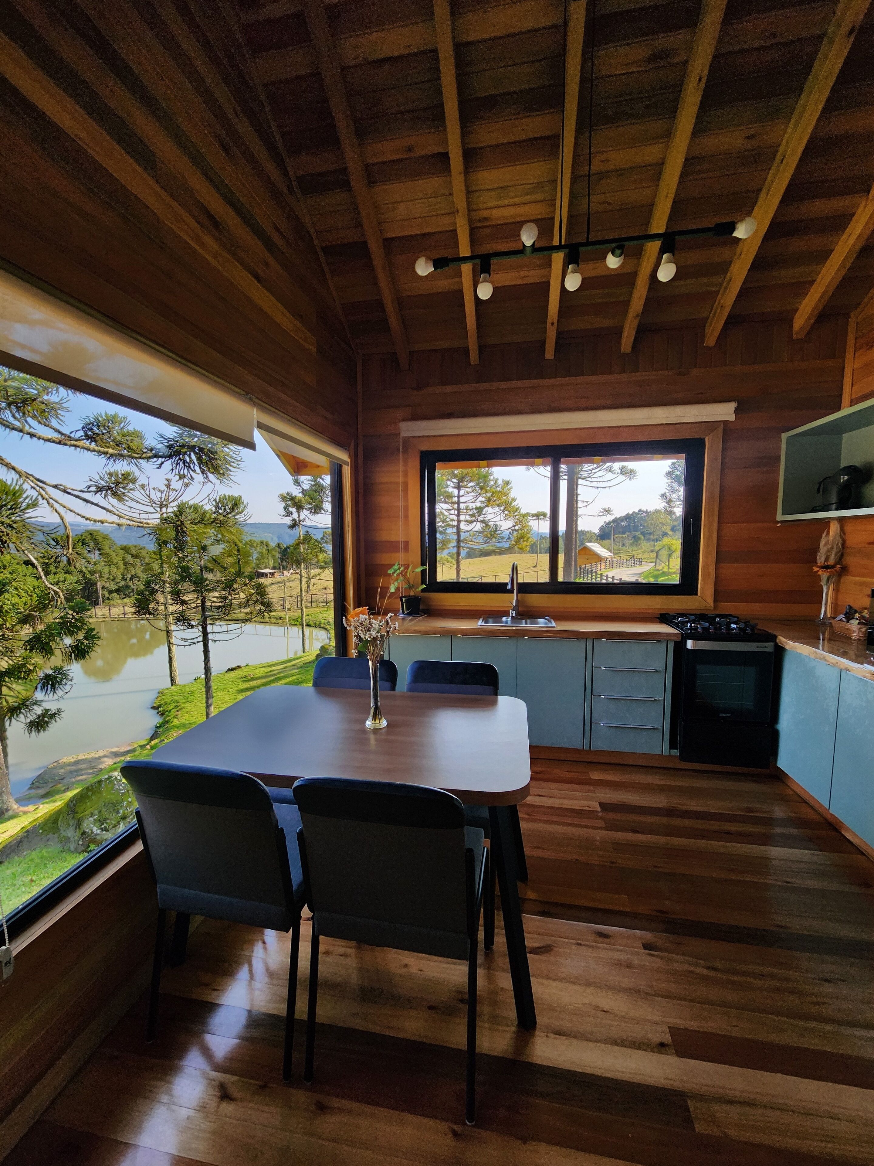 Interior de madeira com mesa de jantar, cadeiras, cozinha e vista para um lago.
