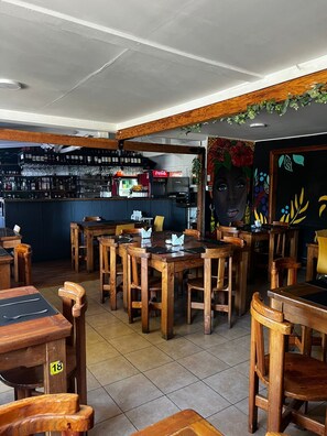 Interior de restaurante com mesas e cadeiras de madeira, área de bar com prateleiras cheias de garrafas e copos, e um mural na parede.