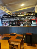 Uma área de bar com prateleiras de garrafas e copos, uma mesa de madeira com cadeiras e um refrigerador.