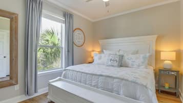 Quarto com uma cama grande, um ventilador de teto, uma janela com cortinas, uma mesa de cabeceira com abajur e um enfeite na parede.