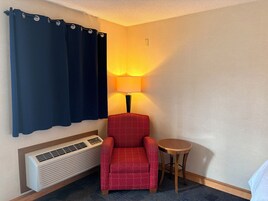 Quarto de hotel com poltrona vermelha, mesa de madeira pequena, aquecedor e luminária.