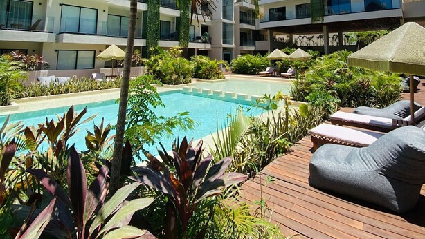 Uma área de piscina com espreguiçadeiras, uma palmeira e um edifício moderno com varandas.