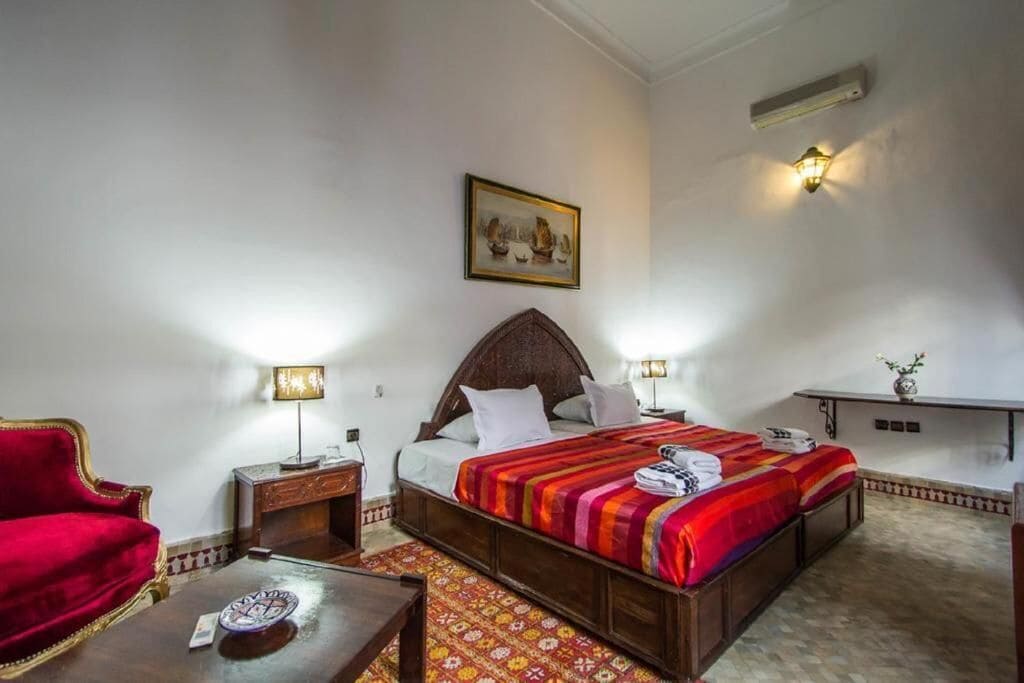 Quarto de hotel com cama, uma poltrona vermelha, uma mesinha, mesa de cabeceira, prateleira na parede e um quadro.