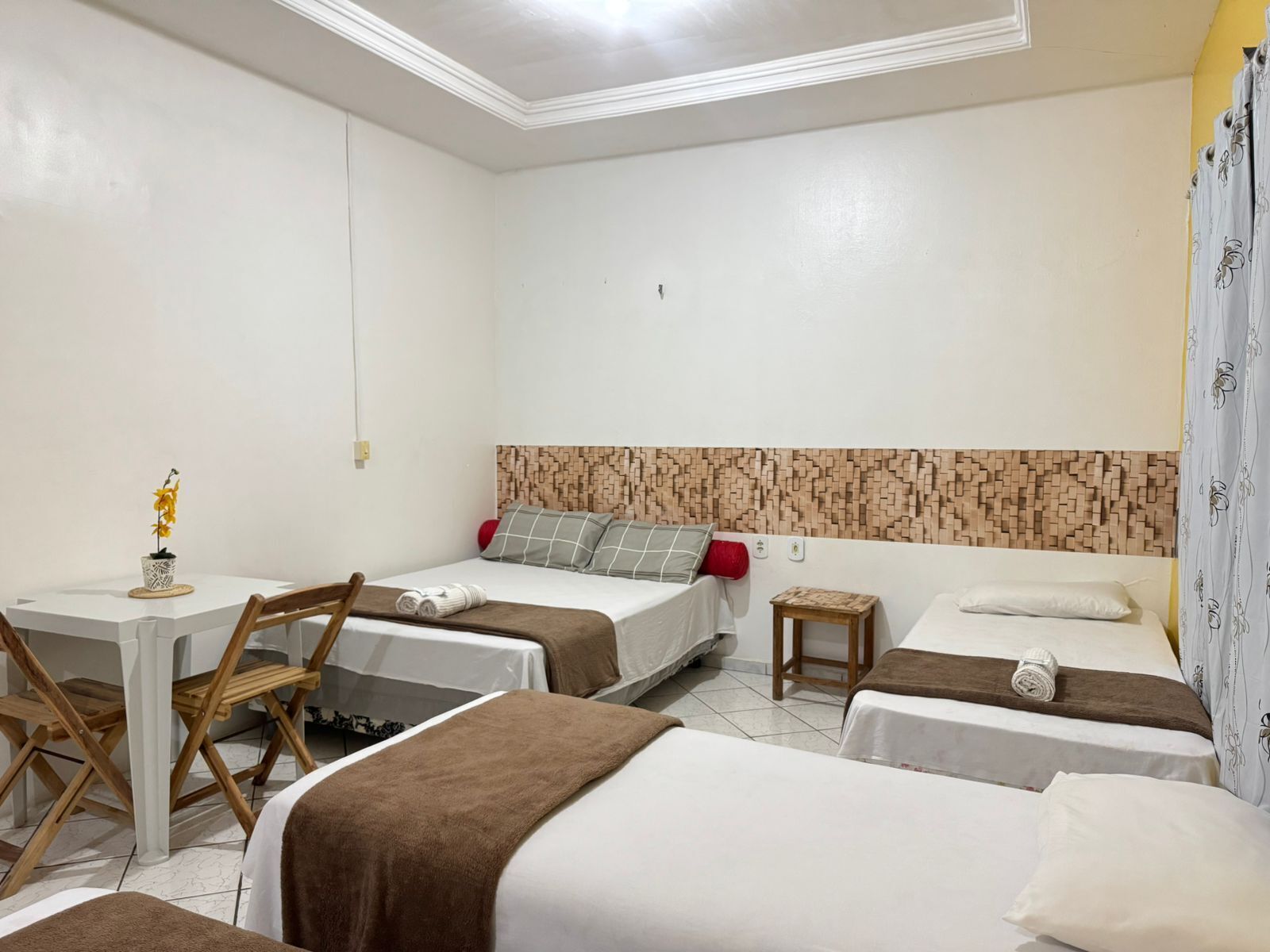 Quarto de hotel com três camas, uma mesinha, uma cadeira e um painel decorativo na parede.