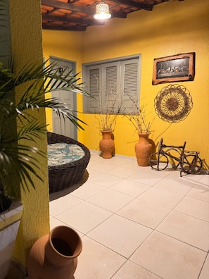 Quarto com paredes amarelas, uma cadeira de vime, vasos de terracota, uma bicicleta e um quadro emoldurado.